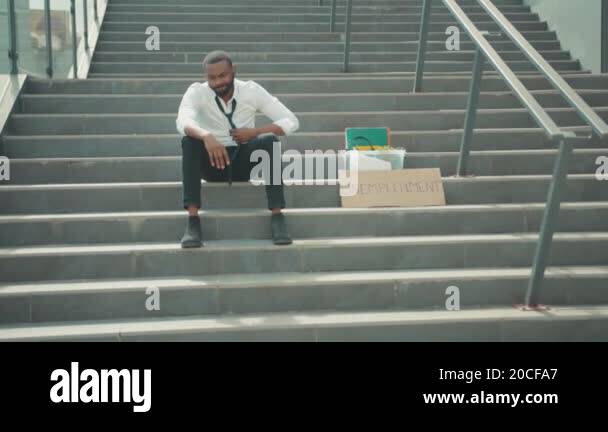 Angry African american young man sitting on stairs with box personal ...