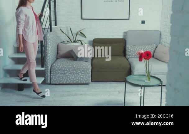 Caucasian young woman walks downstairs into cozy lounge zone living room and starts working on ...