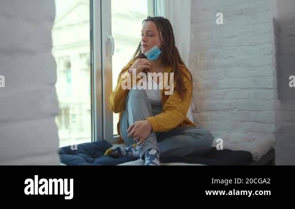 Unhappy young woman sits on windowsill wearing medical face mask self ...