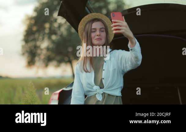Happy young woman with hat speak on smart phone use webcamera in stylish car trunk at sunlight ...
