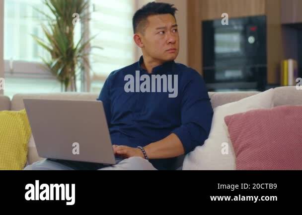 Face serious asian man sitting on sofa use laptop computer work at home ...