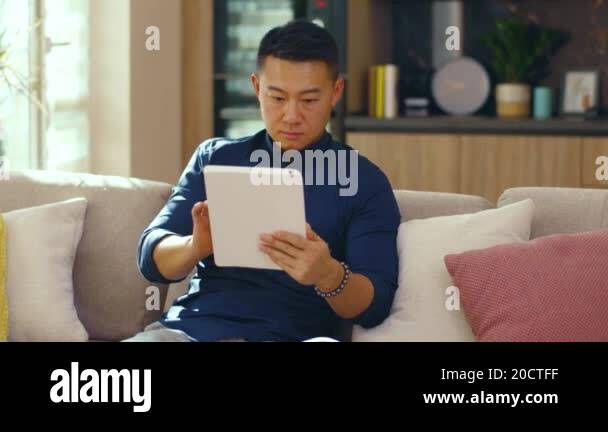 Attractive asian young man use tablet computer sitting on sofa at home ...