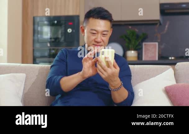 Smiling happy young asian man use phone sitting on sofa at home ...
