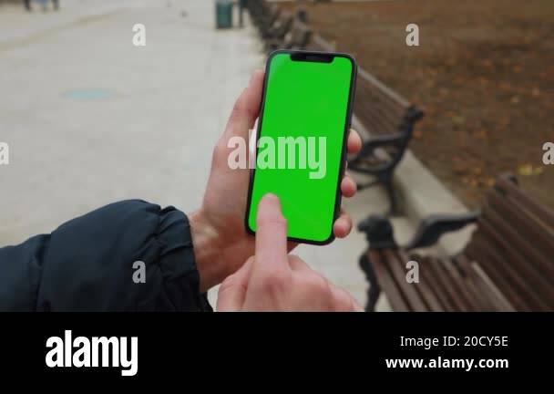 Close up hands man holding use phone with vertical green screen in park background bench ...