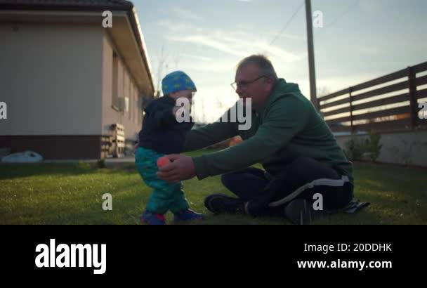 Father sitting on grass helping toddler regain balance after falling on ...