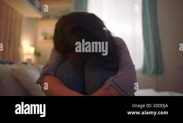 Woman sitting on bed, hugging her knees tightly, head resting forward ...