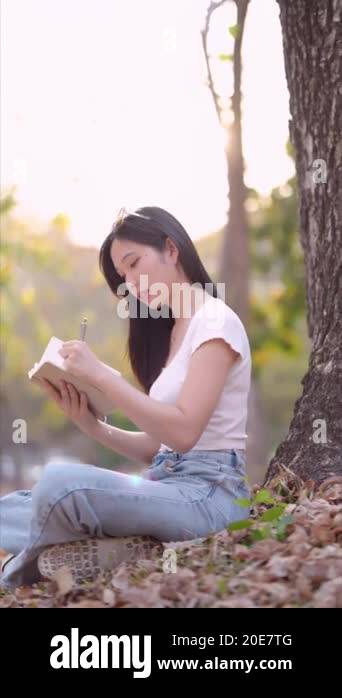 Asian woman capturing thoughts in her journal, finding solace amidst nature at the public park ...