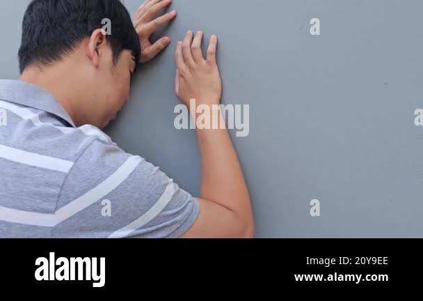 Asian man leans his head against a wall with his hands in despair ...