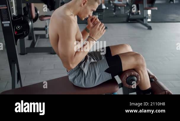 A focused young man sits alone on a bench in a modern gym, contemplating his next move in a ...