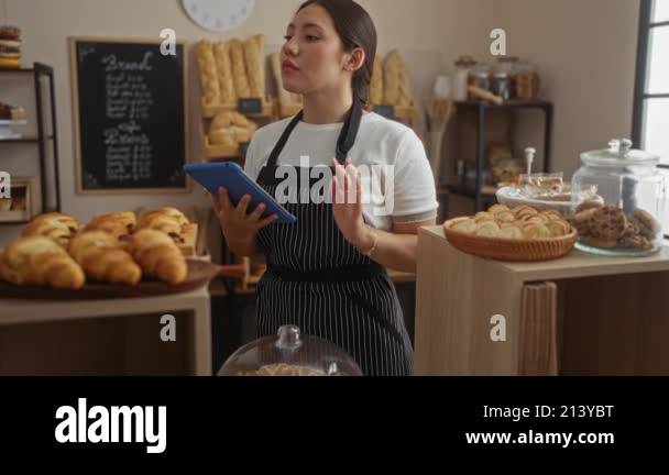 Young woman working in a bakery using a tablet surrounded by bread and ...