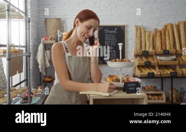 Redhead woman talking on phone and writing in notebook in bakery shop ...