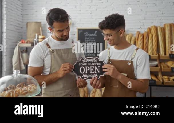 Men bakers holding an 'open' sign in a cozy bakery with bread loaves in ...