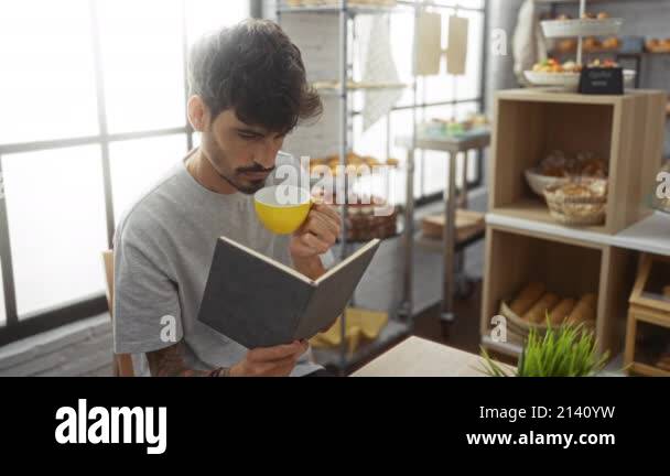 Young man drinking coffee and reading a book in a cozy bakery with ...