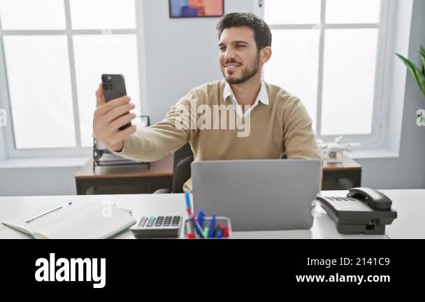 A handsome hispanic man takes a selfie at his office desk with a laptop ...
