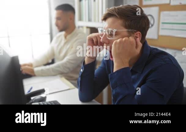 Two men working indoors in an office with one man appearing stressed and another man focused on ...
