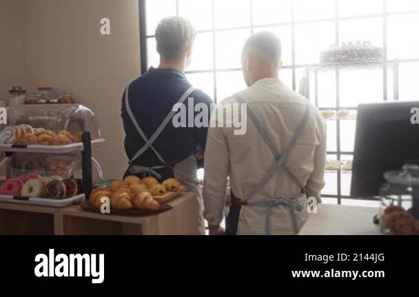 Two smiling male bakers wearing aprons embrace joyfully in a cozy ...