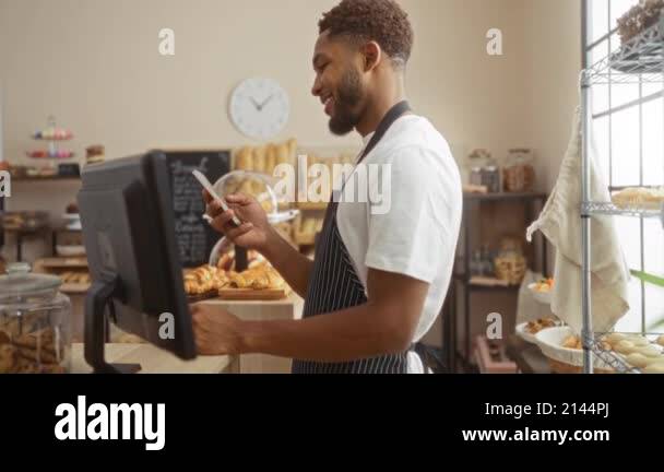 Young man working in a bakery smiling while using a smartphone and ...