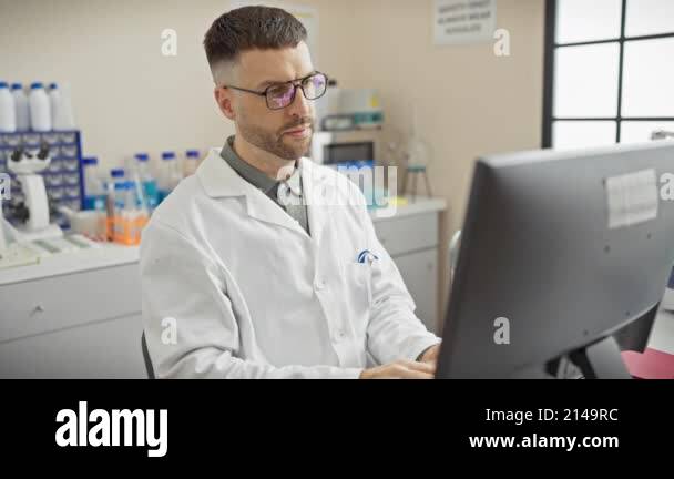 Young adult man with beard in lab coat stretches while removing glasses in laboratory setting ...