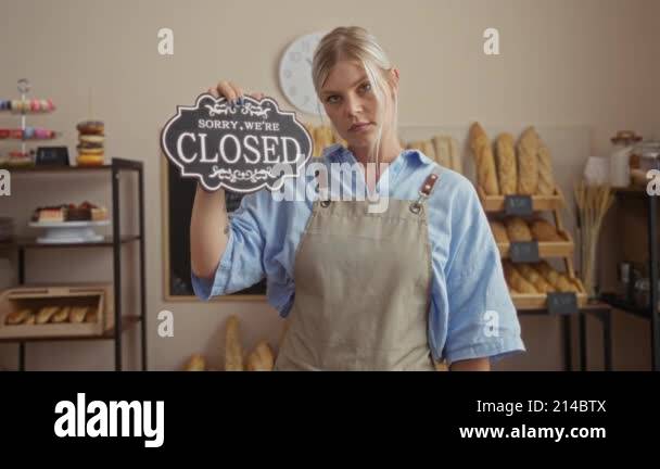 Young woman at bakery holding a closed sign, surrounded by bread and ...