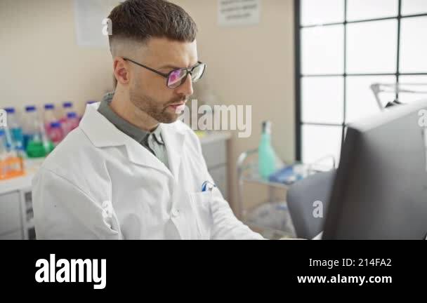 A focused hispanic man in a lab coat analyzes data on a computer screen ...