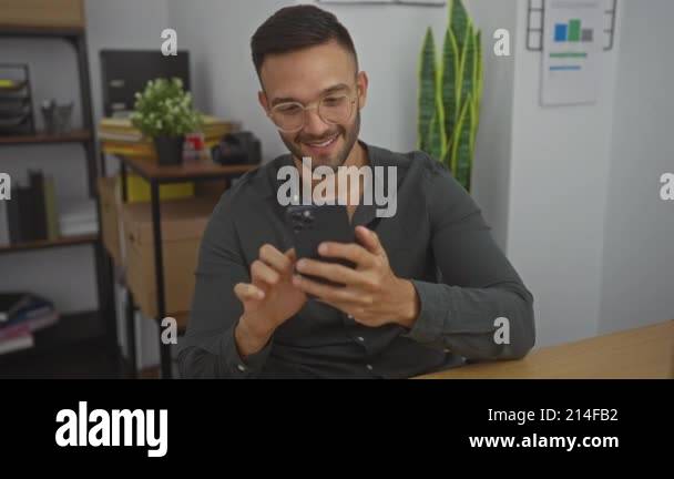 Handsome young hispanic man with a beard smiling and using his smartphone while working in a ...