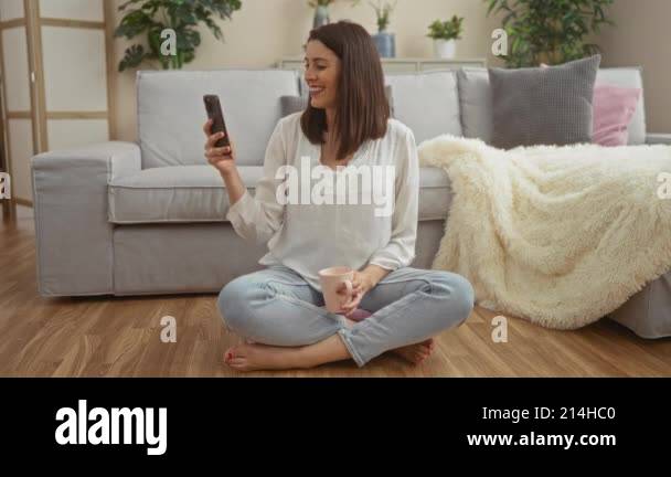 Smiling woman sitting cross-legged on a living room floor with her dog, holding a phone and a ...
