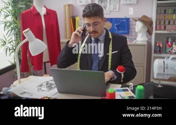 A focused tailor man multitasks with a phone call and laptop in a ...
