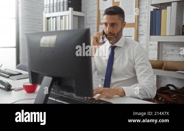 Man in a modern office speaks on the phone while working on a computer, surrounded by ...