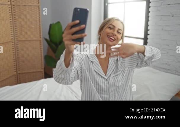 Woman videoconferencing with smartphone in cozy bedroom wearing pajamas ...