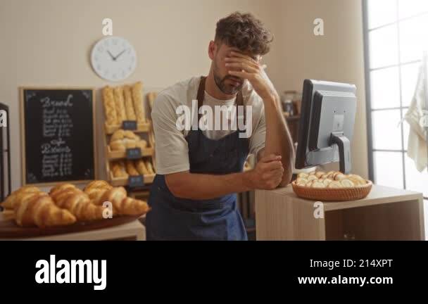 Young man in a bakery wearing an apron leaning on the counter looking ...
