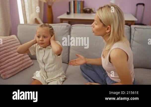 Mother talking to her daughter in the living room, with the child covering her ears, showing a ...