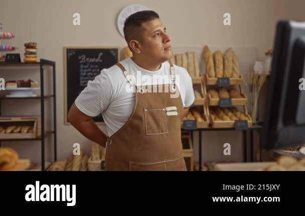 Man working at a bakery wearing a brown apron, surrounded by shelves of bread and pastries, with ...