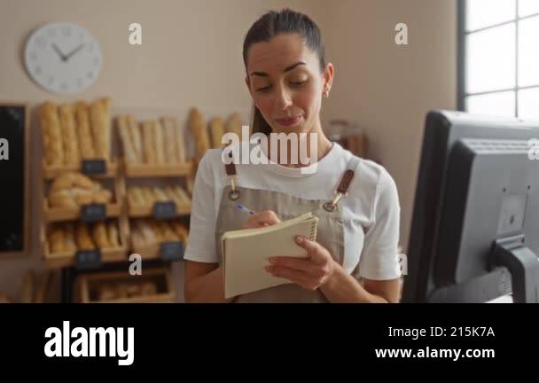 Young woman working in a bakery, writing on a notepad while managing ...