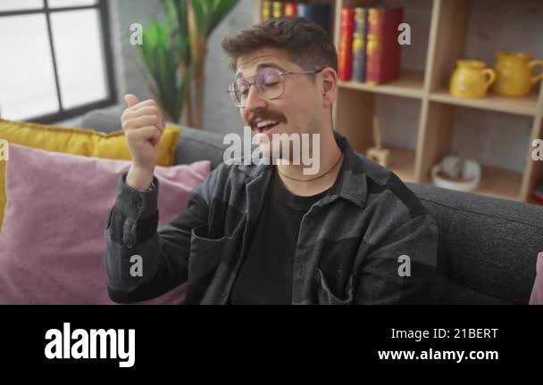 Cheerful young hispanic man with glasses chillin' on the sofa, pointing ...