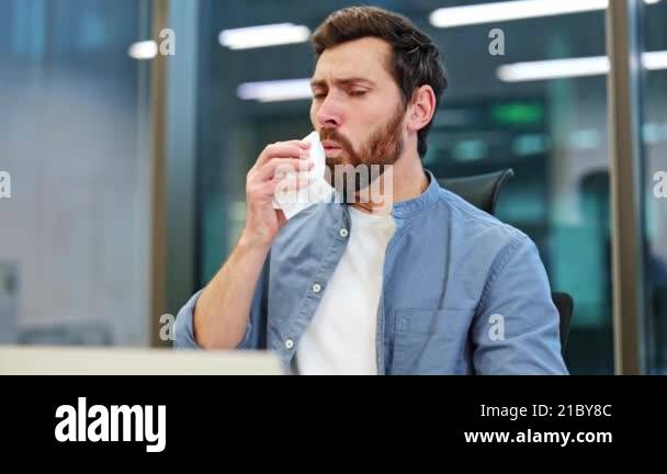 Unwell businessman blowing nose and coughing at work desk in modern ...