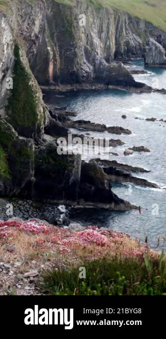 A high-angle view shows a dramatic coastline. Dark cliffs meet the sea ...