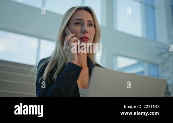 Serious blonde talking cellphone sitting office staircase with laptop ...