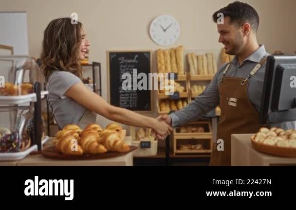 Man and woman smiling and shaking hands in bakery shop interior surrounded by bread and pastries ...