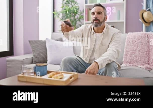 A mature man with a beard and grey hair sits on a sofa in a living room ...