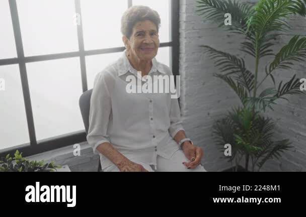 Smiling elderly hispanic woman in white shirt sitting indoors by a ...