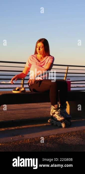 vertical Fit woman preparing to inline skate, sitting on seaside bench during golden sunset ...