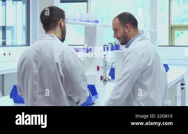 Two male scientists in white lab coats discuss a research project, occasionally joined by a ...