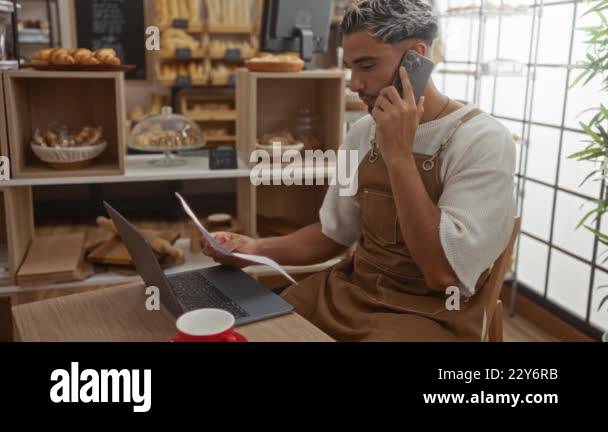 Young man working at bakery using laptop speaking on phone reviewing ...