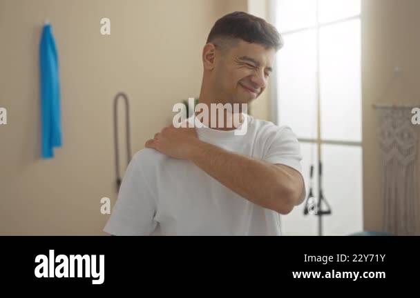 Young man smiling while touching shoulder in gym with bright window and ...