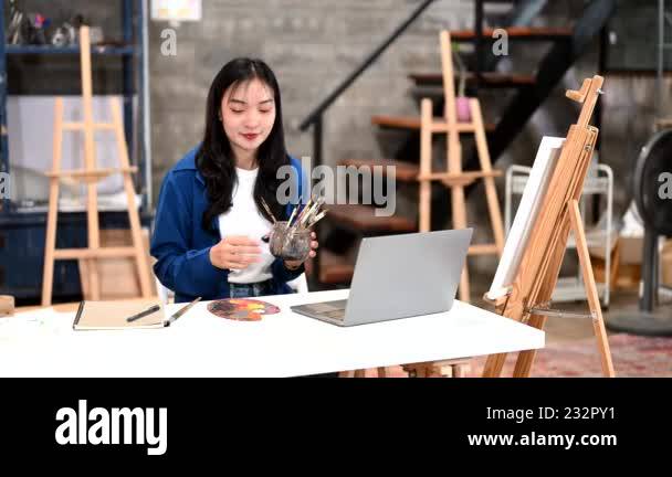 Cheerful female Asian artist using laptop in a vibrant studio with ...