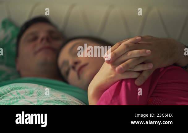 Tender moment of a couple lying closely together in bed, their hands intertwined, conveying ...