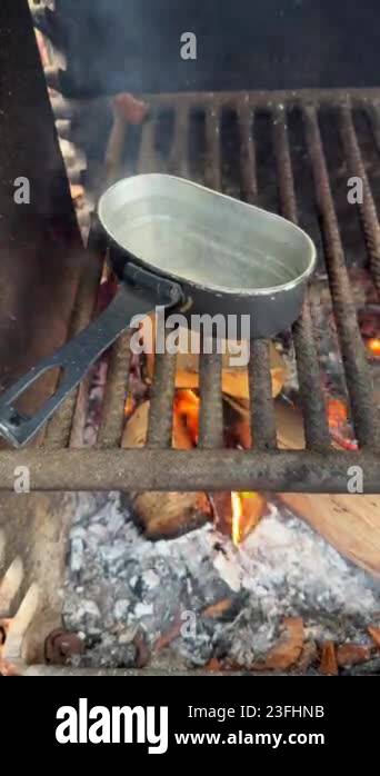 A metal pot sits on a grill over a campfire, with steam rising from the ...
