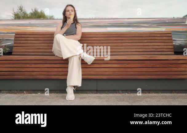 A young woman sits alone on a park bench with her legs crossed, eating ...