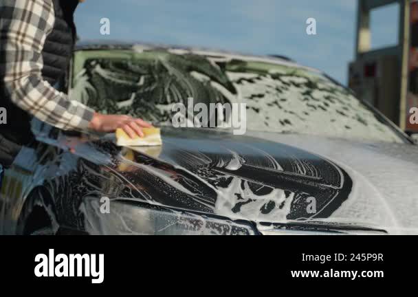 A close-up of a man using a yellow sponge to scrub the hood of a foam ...