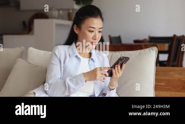 Serious relaxed young Asian cellphone user woman browsing Internet on mobile phone, getting ...